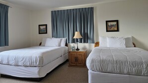 Quarto de hotel com duas camas, um ventilador de teto, mesa de cabeceira com abajur e duas quadros nas paredes.
