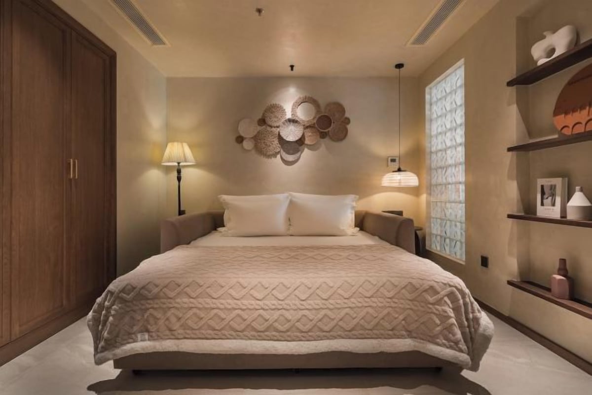 Quarto com uma cama grande, duas luminárias de cabeceira, um objeto decorativo na parede e estantes embutidas com objetos decorativos.
