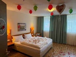 Quarto de hotel com cama, mesa de cabeceira, luminária, quadro e janela com cortinas.