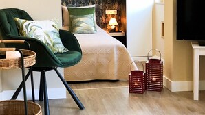 Quarto moderno com uma cadeira verde, cama com almofadas estampadas, mesa de cabeceira com abajur, televisão e cestos de vime.