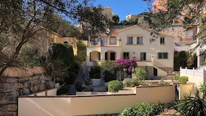 Uma villa em estilo mediterrâneo com fachada de pedra, várias varandas e um jardim paisagístico com escadaria para a entrada.
