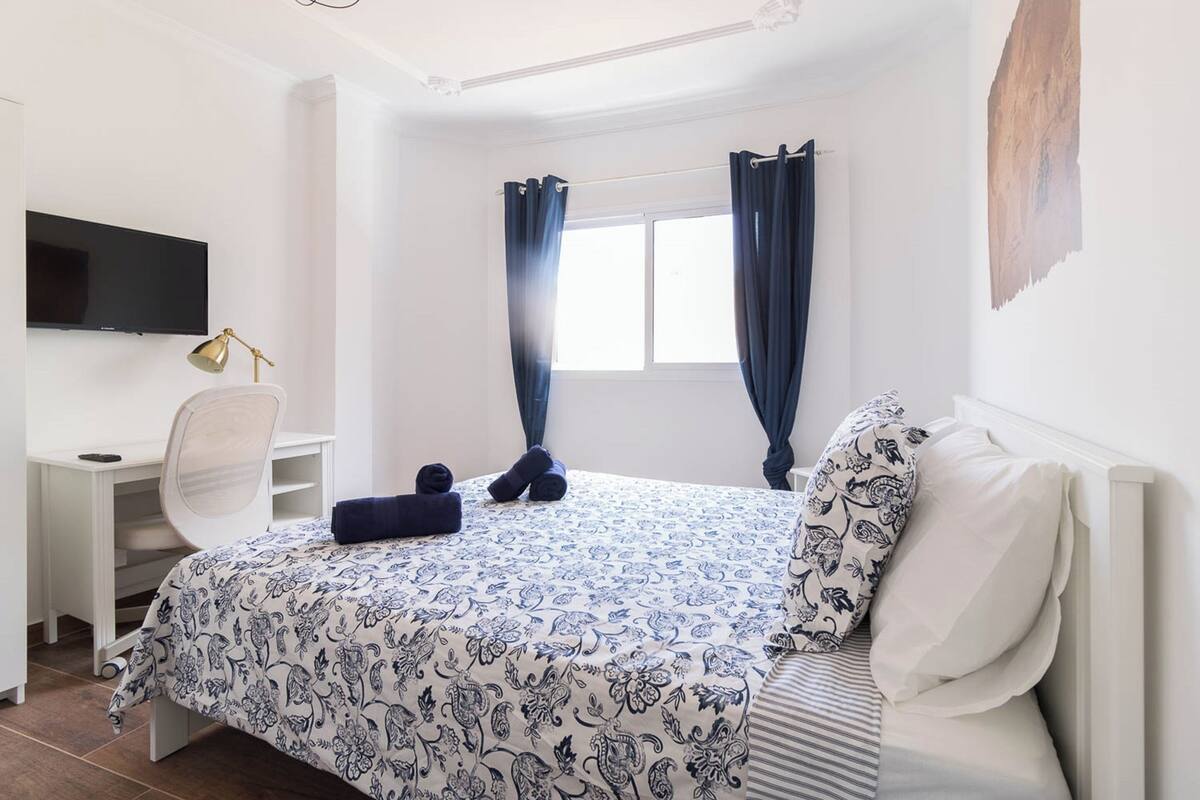 Quarto com cama, escrivaninha, cadeira, televisão e janela com cortinas.