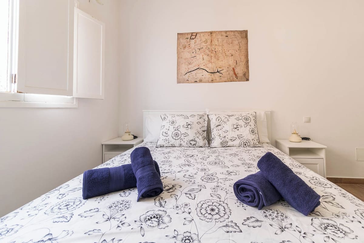 Uma cama bem arrumada com roupa de cama floral, duas toalhas enroladas, uma obra de arte de madeira na parede e uma janela com persianas brancas.
