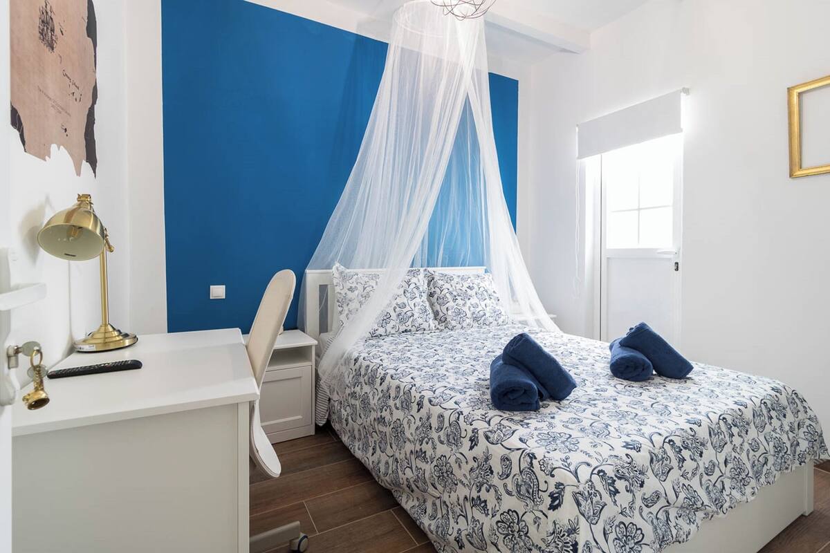 Quarto com cama sob um dossel branco, mesa de cabeceira com abajur e parede azul.