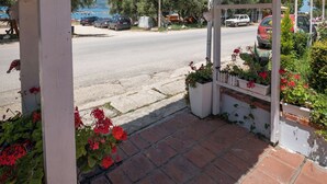 Área externa coberta com telhado de palha, plantas em vasos e vista para uma estrada e carros estacionados.