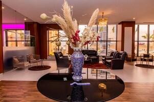 Um lobby moderno com uma mesa redonda central, um vaso decorativo com flores, sofás de couro e grandes janelas com vista para a cidade.