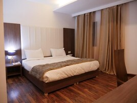 Quarto de hotel com uma cama grande, piso de madeira, mesa de cabeceira com abajur e vista para o exterior através de cortinas.