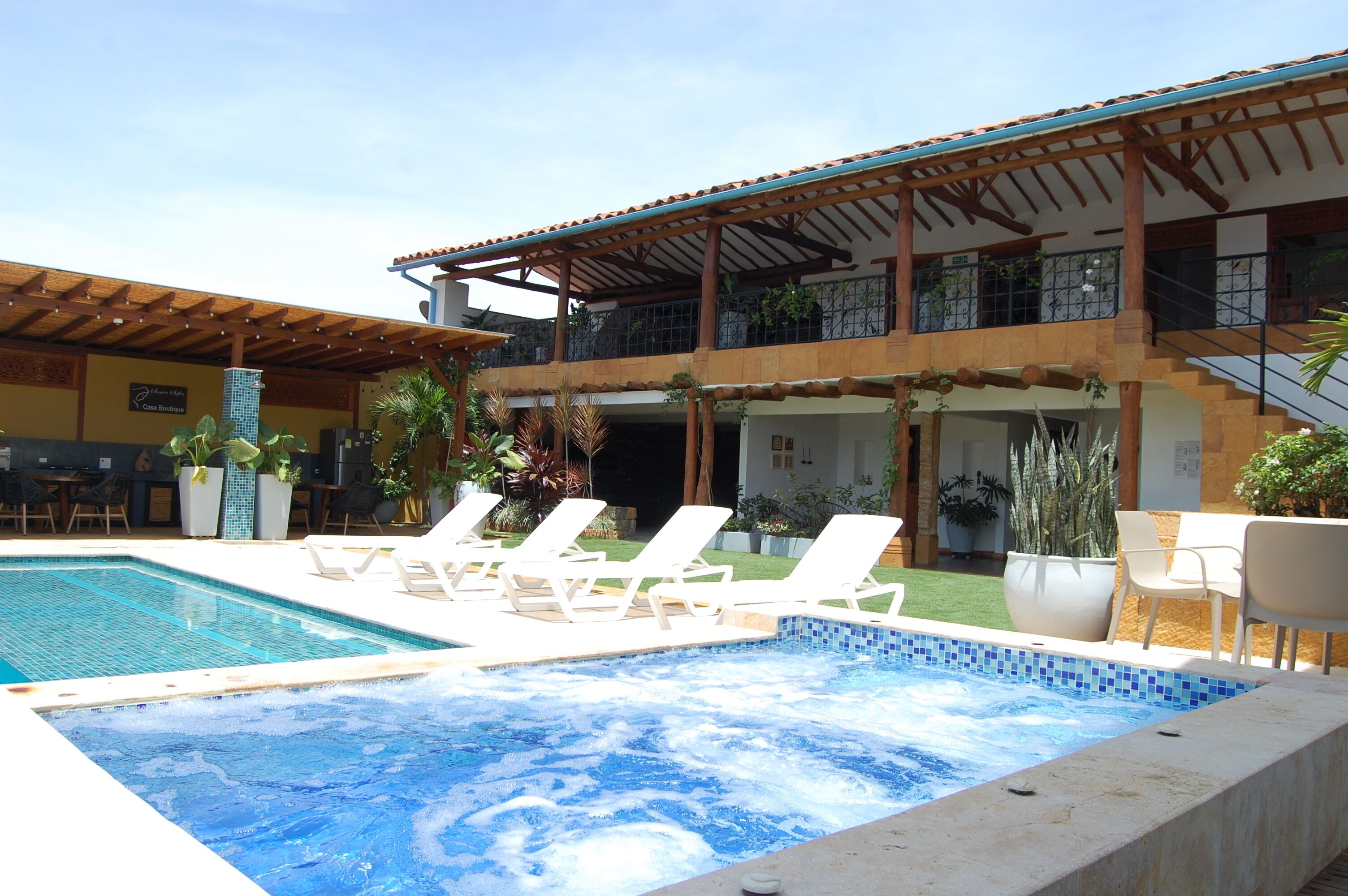 Uma área de piscina com espreguiçadeiras, uma banheira de hidromassagem e um edifício com telhado em azulejos e varandas.