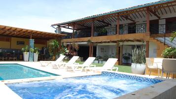 Uma área de piscina com espreguiçadeiras, uma banheira de hidromassagem e um edifício com telhado em azulejos e varandas.