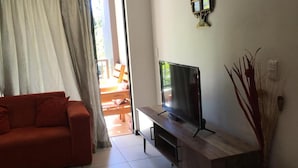 Uma sala de estar com um sofá vermelho, uma televisão de tela plana sobre um suporte de madeira, e vista para o exterior através de uma janela grande.
