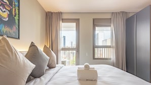Quarto de hotel com uma cama grande, duas janelas com cortinas, vista para prédios e um quadro na parede.