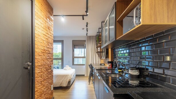 Cozinha moderna com bancadas pretas e um micro-ondas. Uma cama com roupa de cama branca aparece ao fundo.
