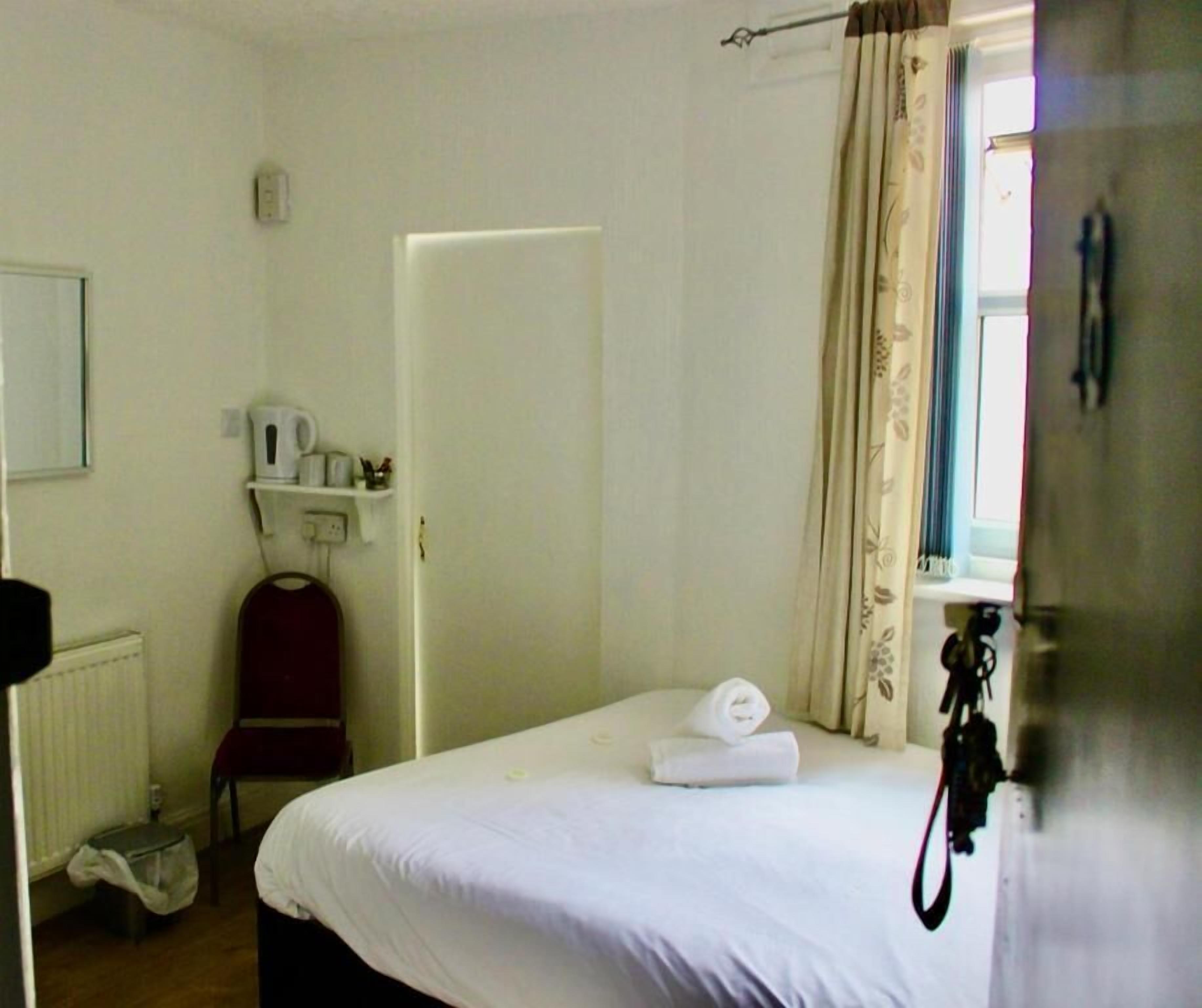 Quarto de hotel com uma cama, um radiador, uma estante com produtos de higiene e uma janela com cortinas.
