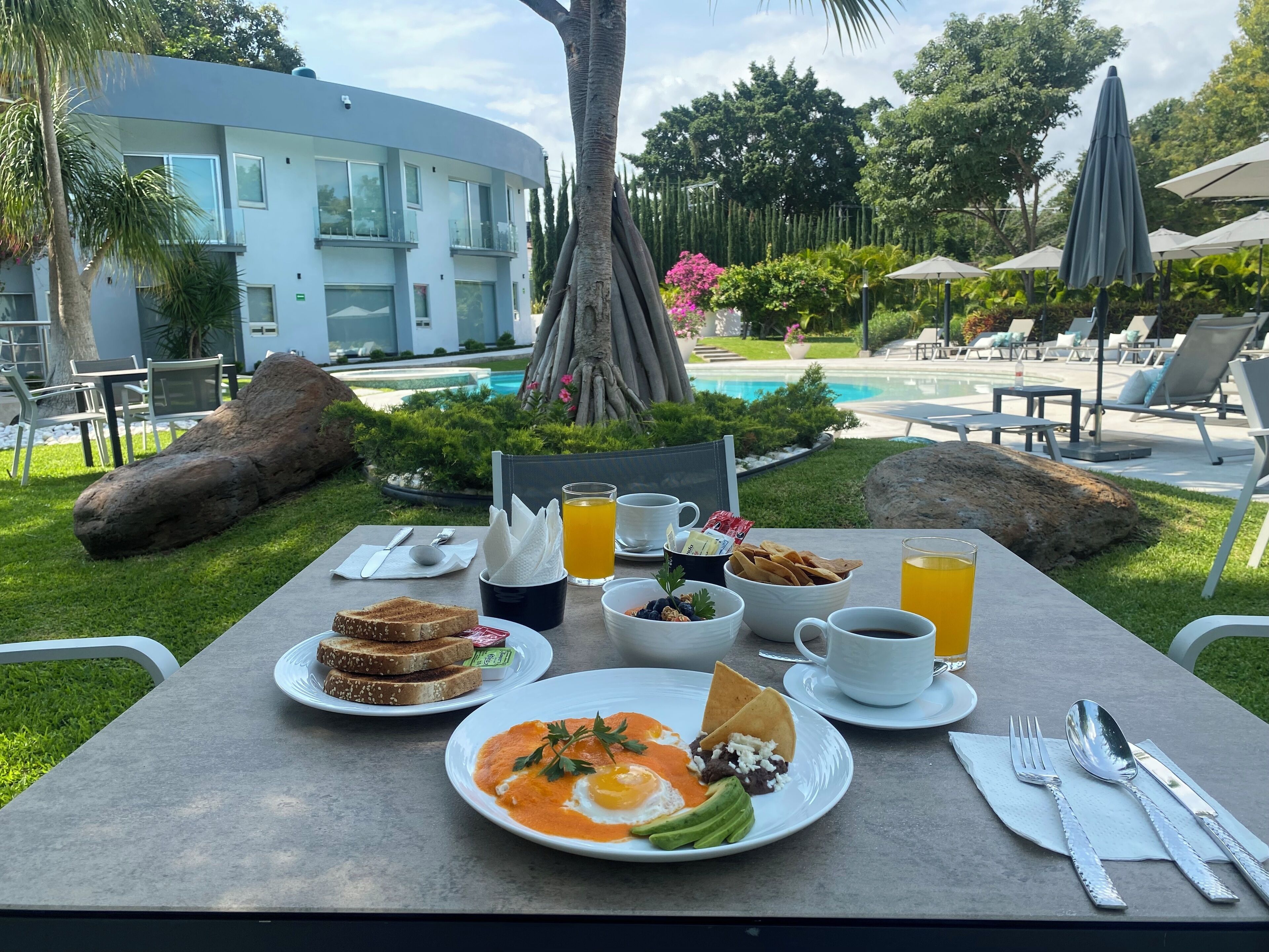 Mesa de jantar com itens de café da manhã, piscina e espreguiçadeiras em um ambiente de resort.