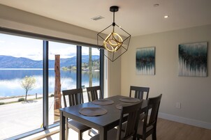 Uma área de refeições com mesa posta para quatro pessoas, janelas amplas com vista para um lago e montanhas, e duas pinturas abstratas na parede.