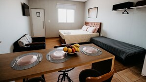 Um quarto pequeno e bem iluminado com uma cama, uma mesa de cabeceira com frutas, uma mesa de jantar com pratos e talheres, e uma televisão na parede.