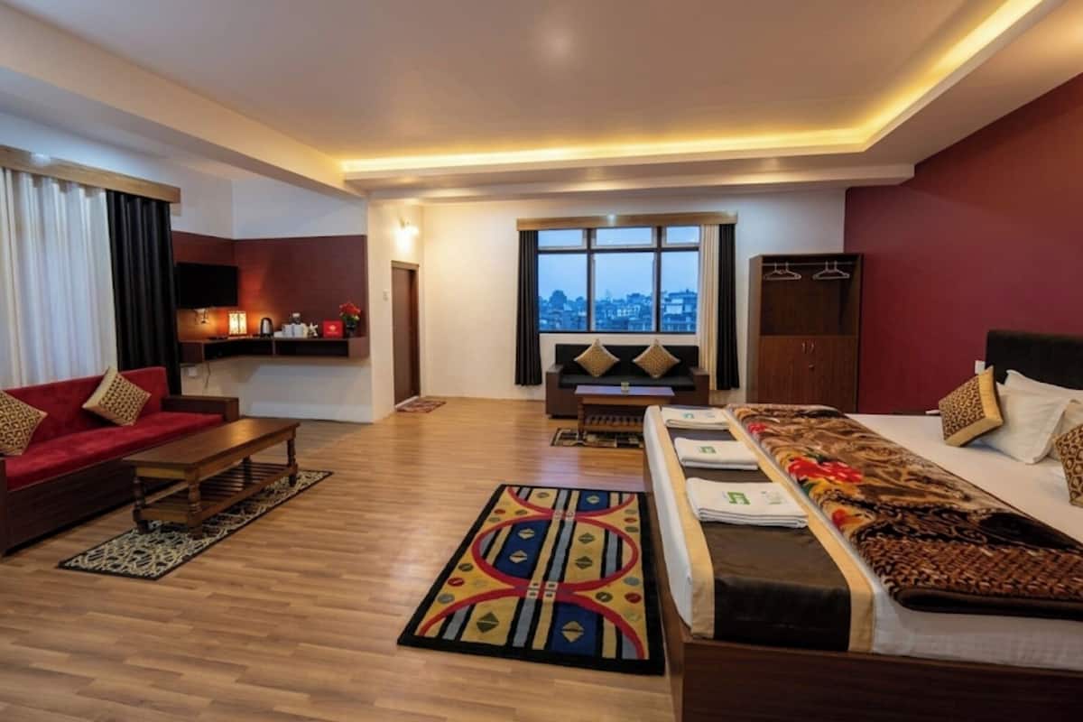Quarto de hotel com cama, sofá, mesa pequena, banqueta, televisão e vista para a cidade.