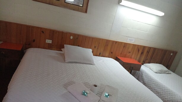 Quarto de hotel com duas camas, cabeceiras de madeira, roupa de cama branca e luminária fixada na parede.