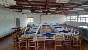 Uma sala de conferências espaçosa com mesas e cadeiras compridas, teto alto com vigas aparentes e amplas janelas de um lado.