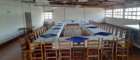 Uma sala de conferências espaçosa com mesas e cadeiras compridas, teto alto com vigas aparentes e amplas janelas de um lado.