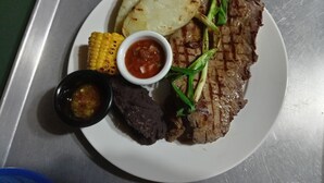 Prato com bife grelhado, espiga de milho, batatas assadas, vagens e dois molhos para acompanhar.
