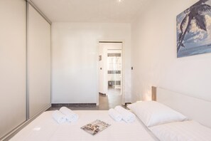 Quarto de hotel com cama, duas toalhas enroladas, uma revista e uma pintura de uma palmeira.