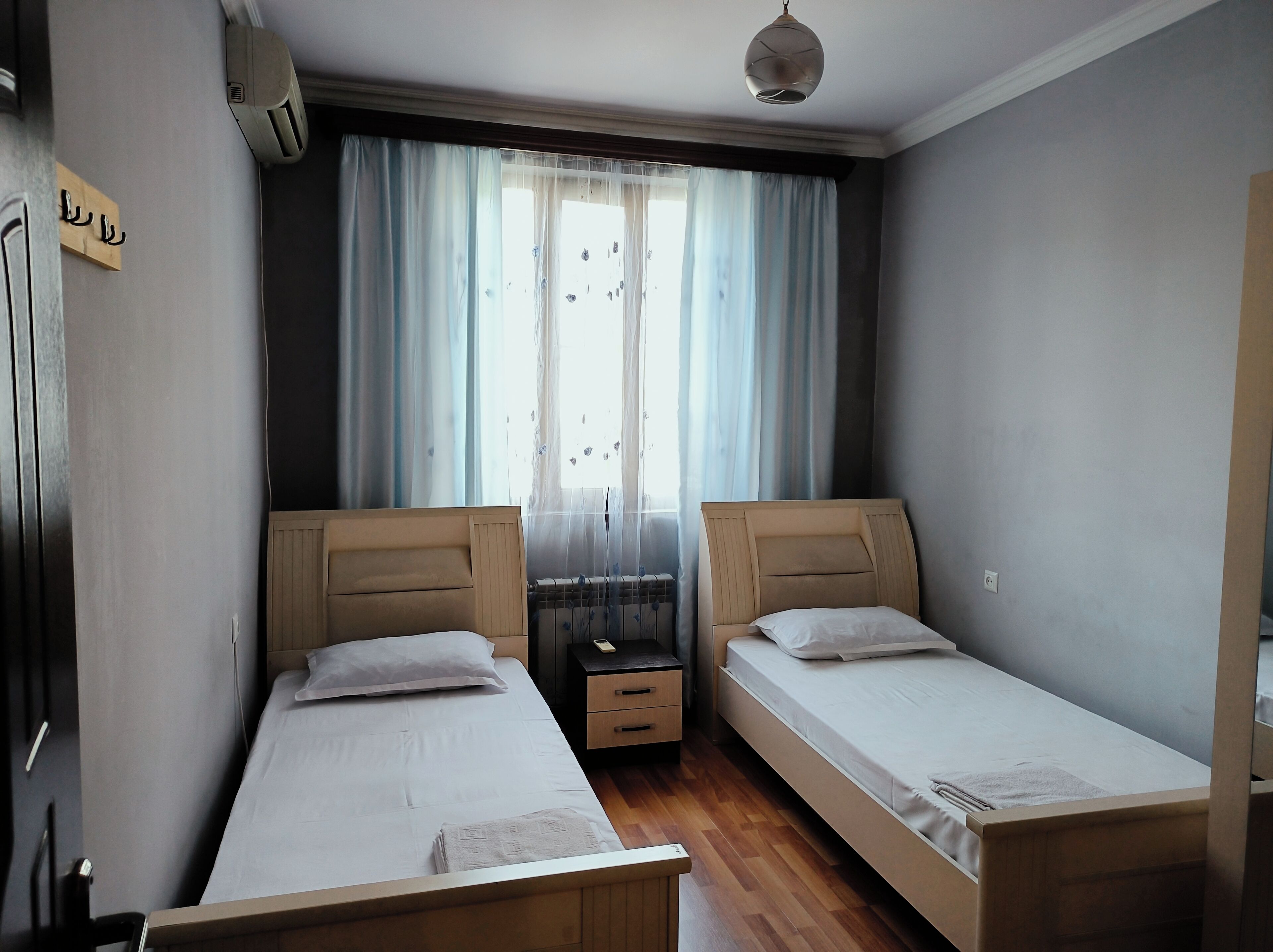 Quarto com duas camas de solteiro, uma pequena mesa de cabeceira, aquecedor e janela com cortinas finas.