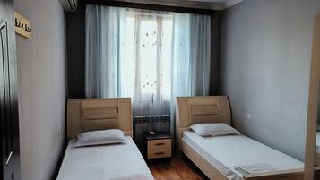 Quarto com duas camas de solteiro, uma pequena mesa de cabeceira, aquecedor e janela com cortinas finas.