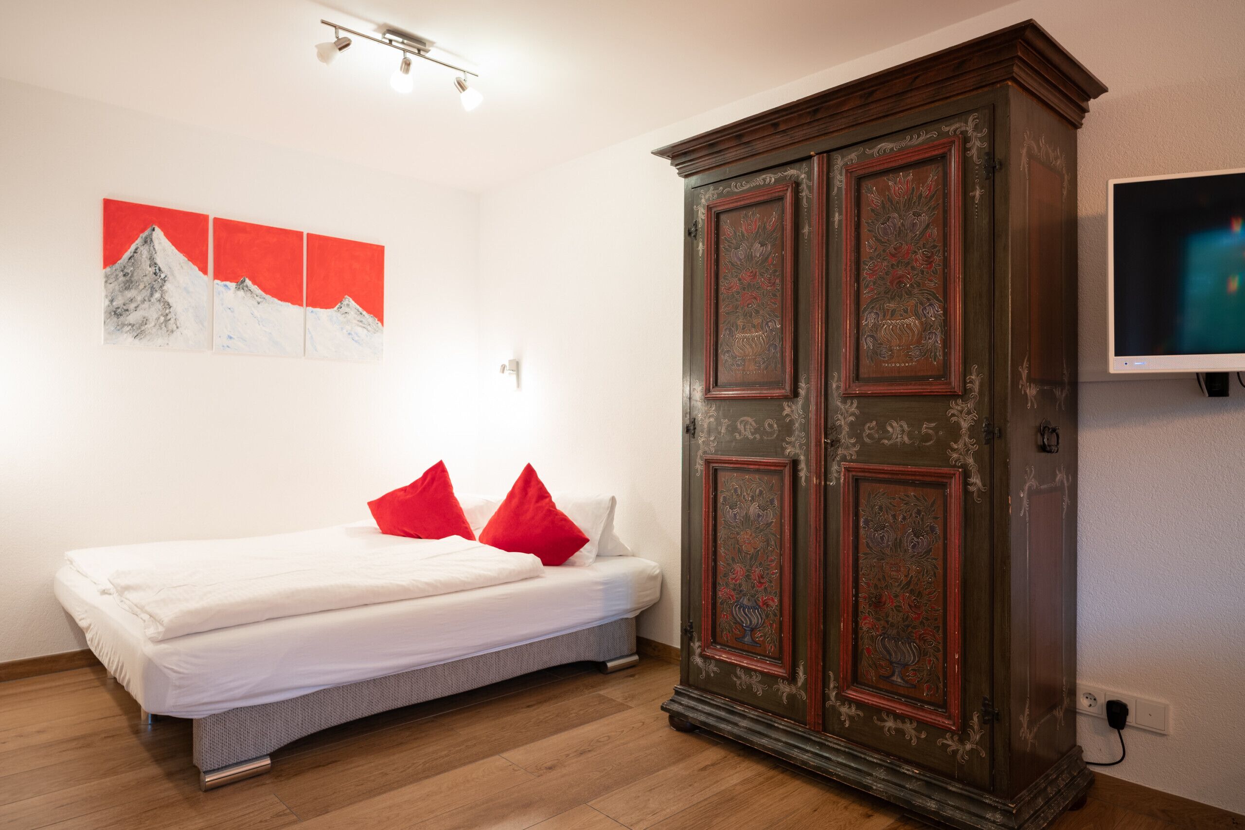 Quarto com cama, almofadas vermelhas, um armário de madeira e uma televisão presa à parede.