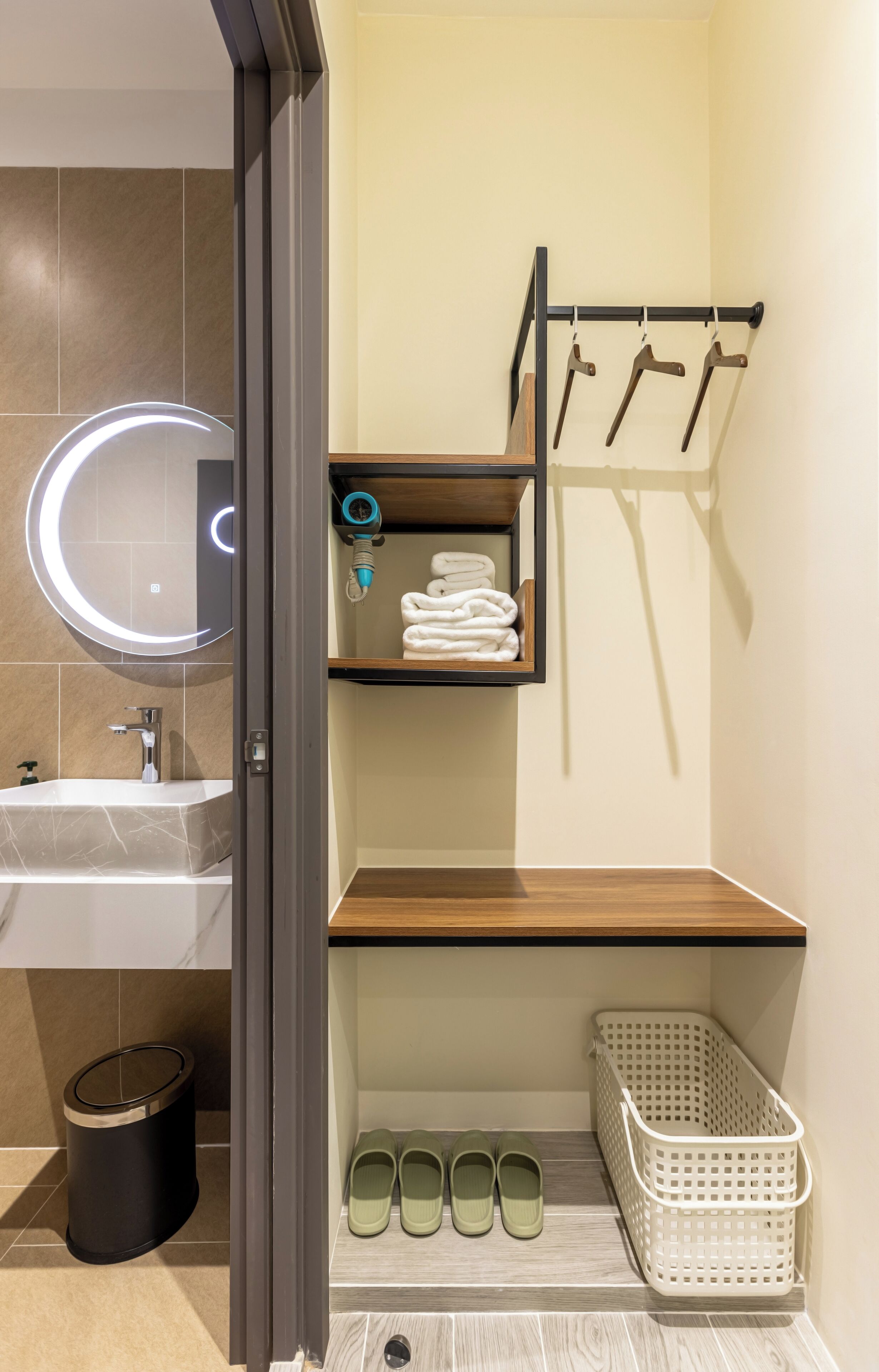 Uma casa de banho com lavatório, espelho iluminado circularmente, porta-toalhas, prateleira com toalhas dobradas, um par de chinelos e um cesto de lixo.