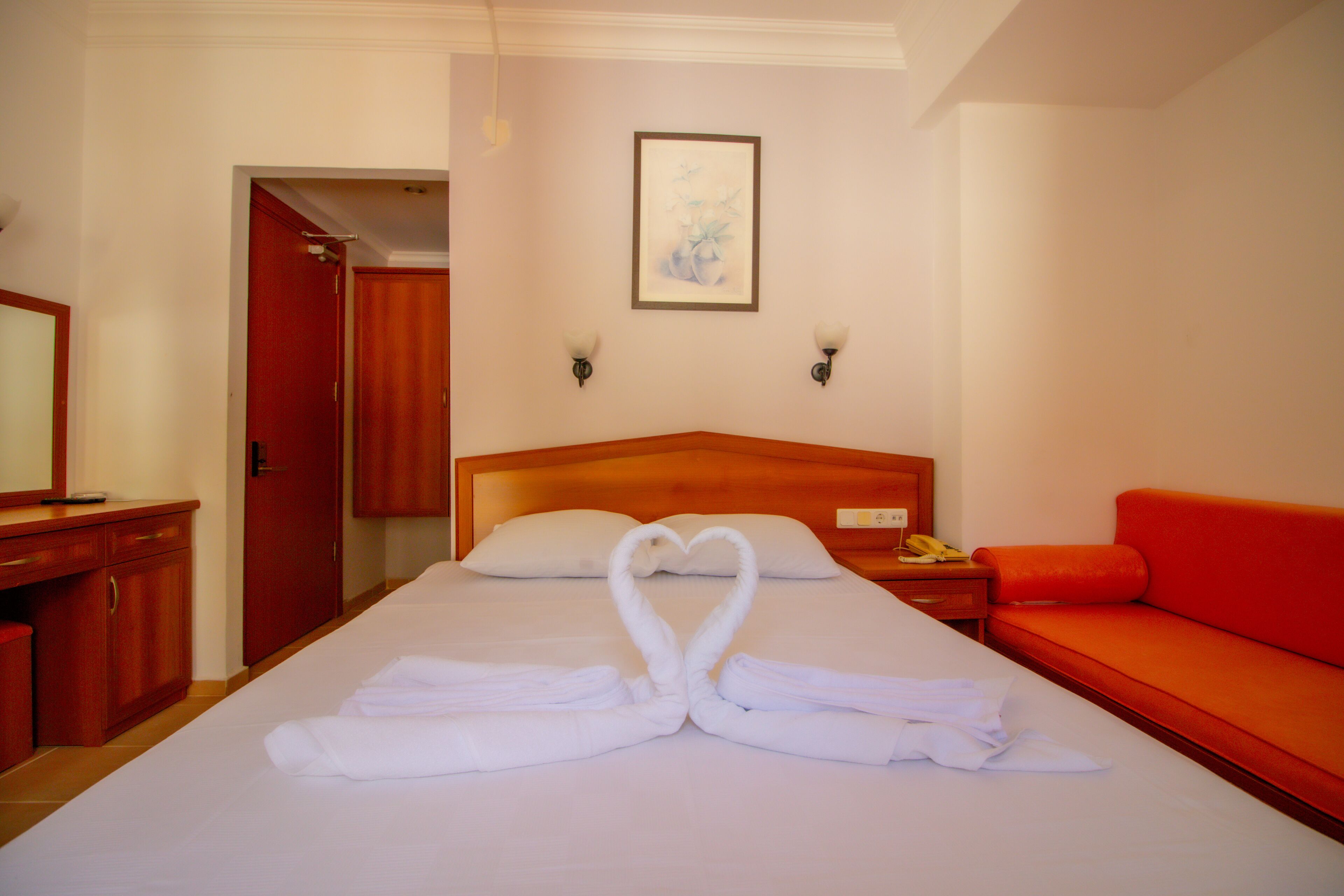 Quarto de hotel com uma cama, um sofá vermelho, uma mesinha de cabeceira de madeira e um quadro na parede.