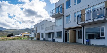 Um edifício moderno de vários andares com amplas janelas e varandas, entrada com calçamento de cascalho e montanhas ao fundo.