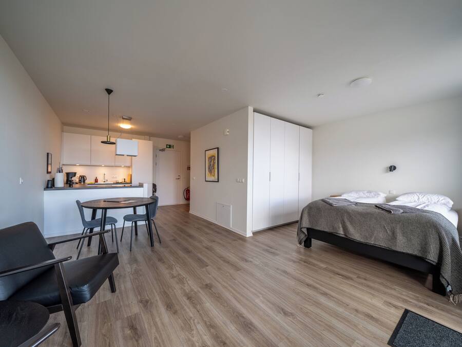 Quarto de hotel moderno com cama, secretária e cozinha compacta.