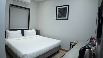 Quarto de hotel com uma cama, uma mesinha com chaleira, um quadro na parede e um ar condicionado sobre a cama.