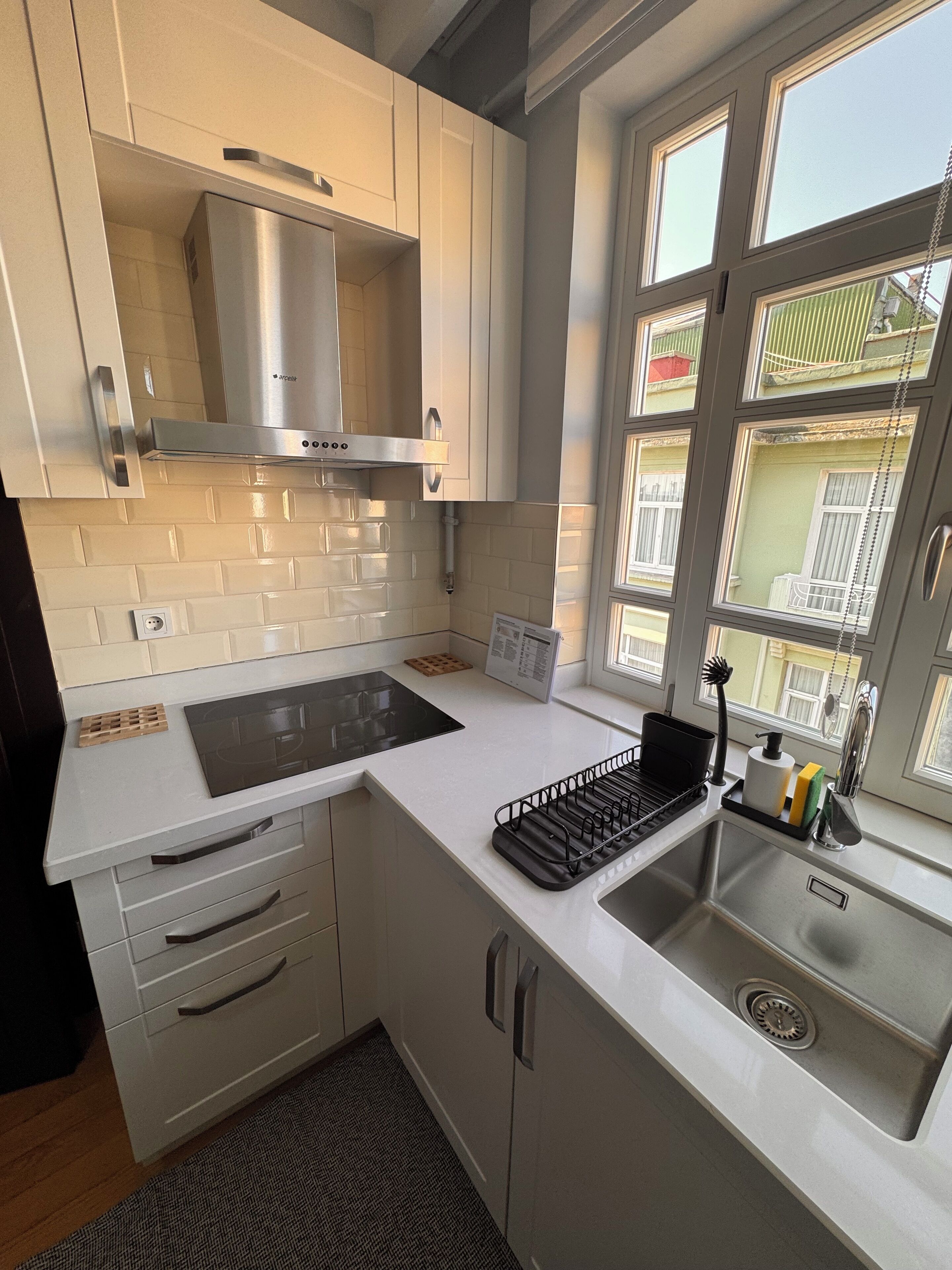 Uma cozinha moderna com armários brancos, um exaustor de aço inoxidável, um fogão por indução preto e uma janela com vista para edifícios.