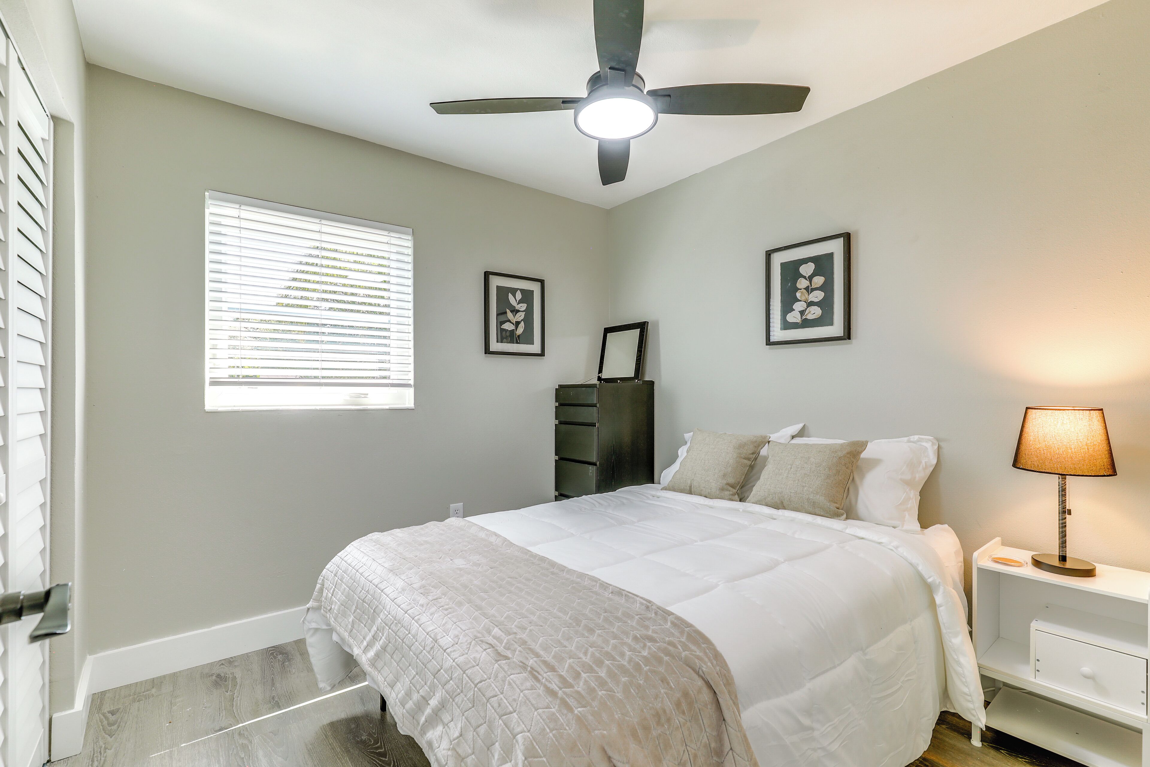 Quarto com cama, ventilador de teto, mesa de cabeceira com abajur, cômoda e duas fotos emolduradas na parede.