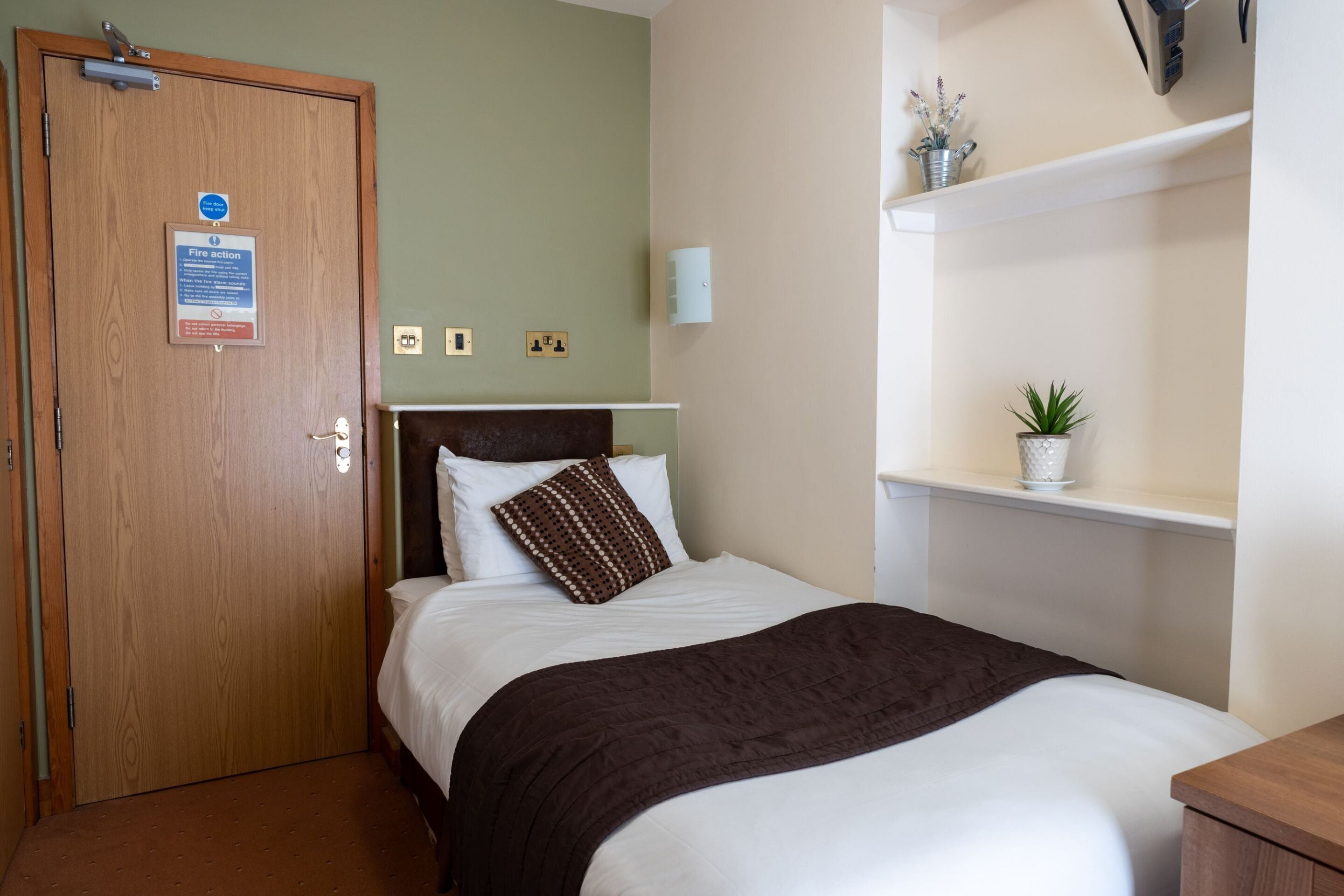 Uma cama bem arrumada com um cobertor marrom e uma almofada decorativa, uma porta de madeira com sinal de emergência contra incêndio e uma pequena prateleira com uma planta.