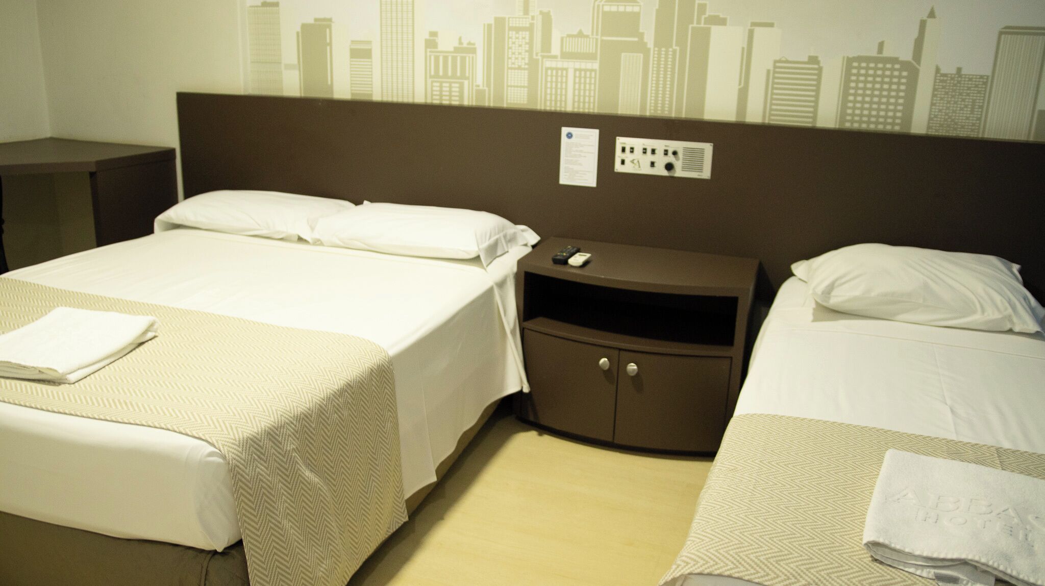 Quarto de hotel com duas camas, uma mesa de cabeceira e uma parede com mural da paisagem urbana.