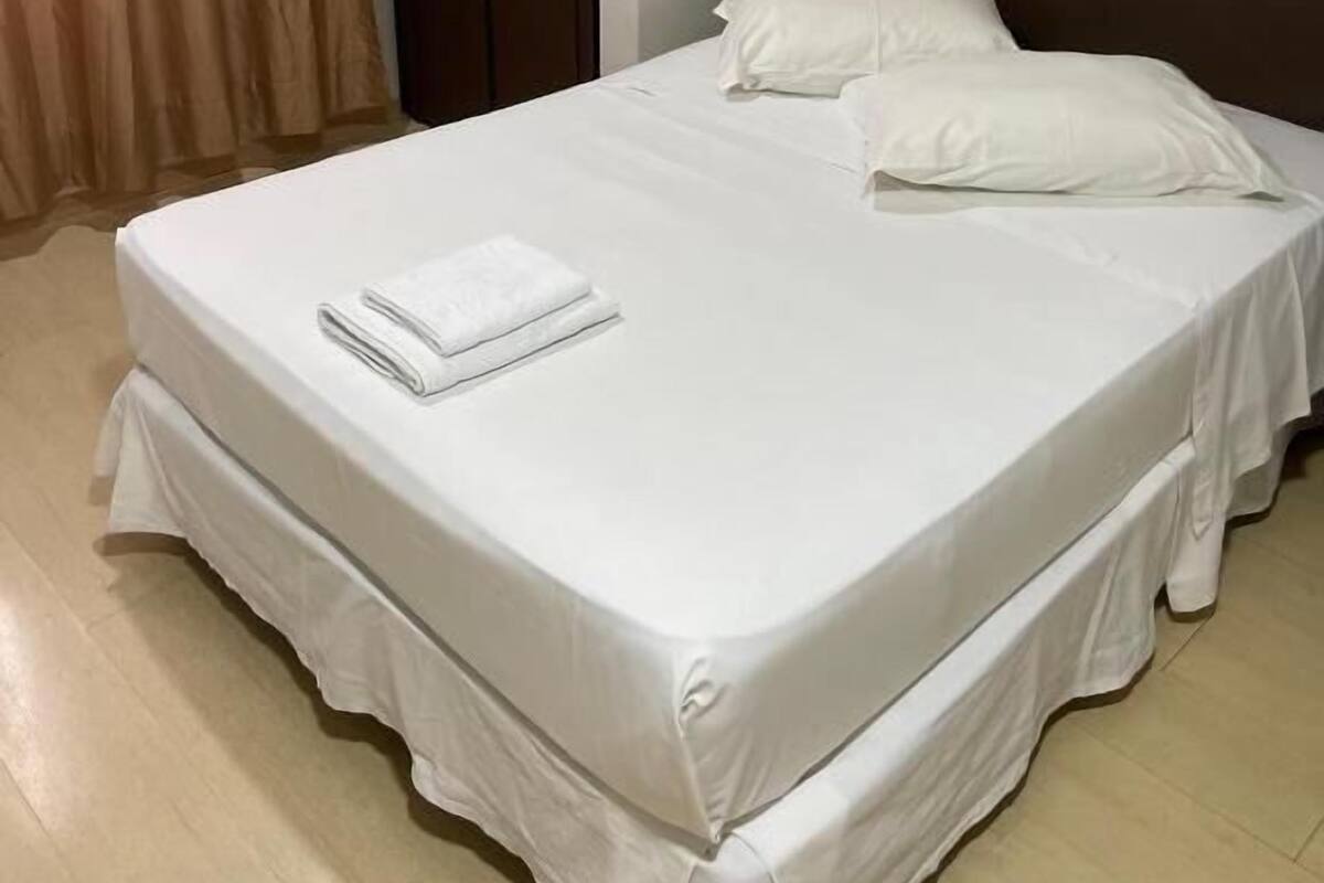 Cama bem arrumada com lençóis brancos e toalha dobrada, guarda-roupa castanho e ar-condicionado fixado na parede.