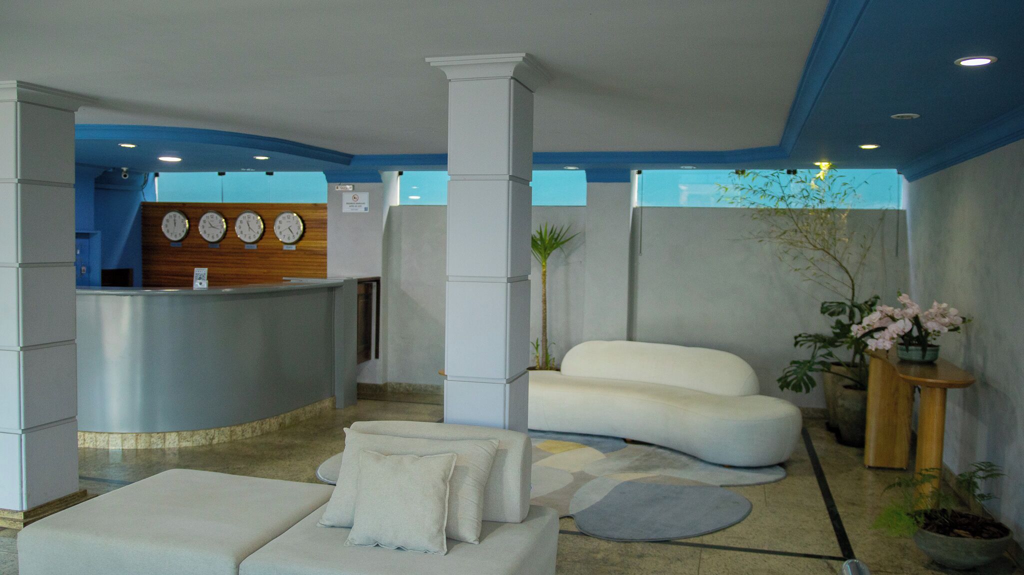 Uma área de recepção com um balcão moderno, espaço de estar com sofá e almofadas, e uma parede de recepção decorada com relógios.