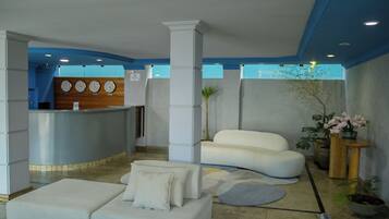 Uma área de recepção com um balcão moderno, espaço de estar com sofá e almofadas, e uma parede de recepção decorada com relógios.