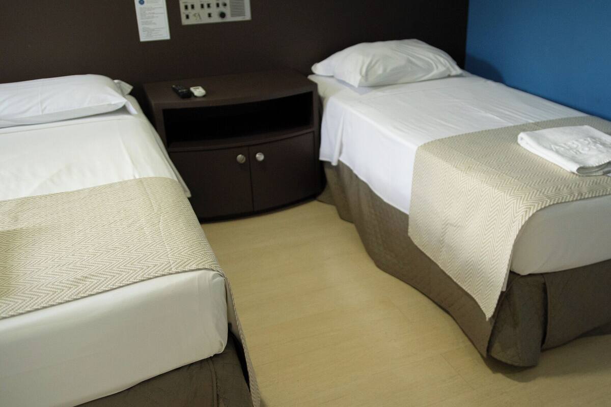 Quarto de hotel com duas camas de solteiro, uma pequena mesa de cabeceira e uma tomada na parede.