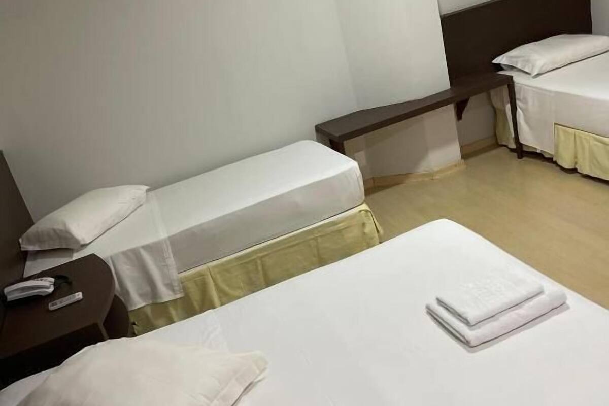 Quarto de hotel com duas camas de solteiro, uma pequena mesa de cabeceira com telefone e controle remoto, e um banco encostado na parede.