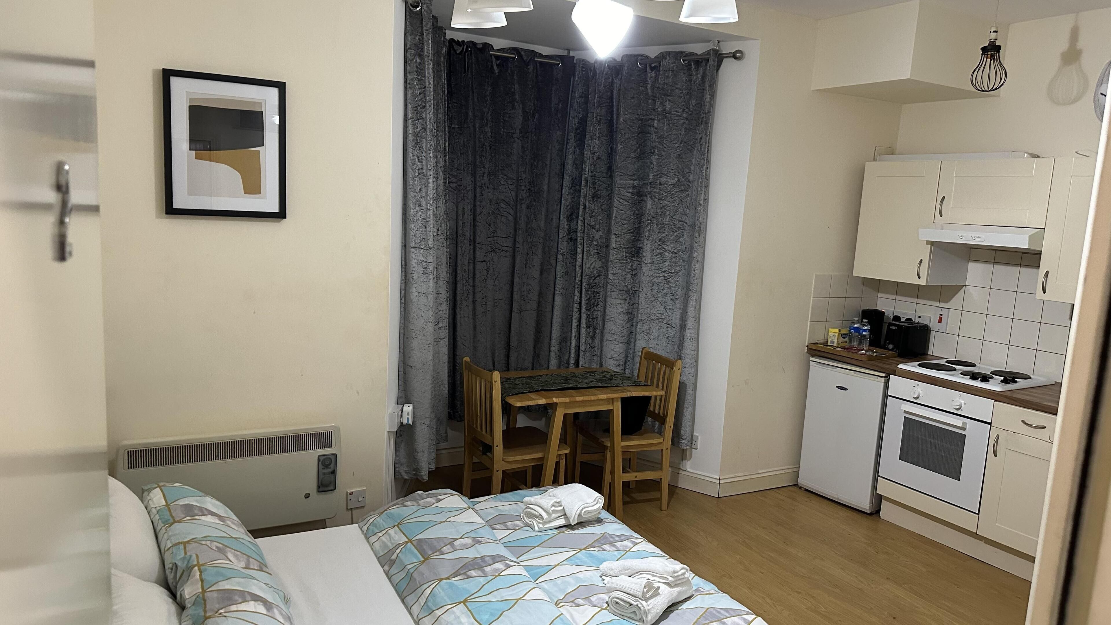 Quarto de hotel compacto com cama, pequena mesa de jantar com cadeiras, cozinha compacta e um único quadro na parede.