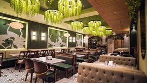Interior de restaurante com paredes verdes, arte nas paredes em branco, plantas pendentes e diferentes opções de assento, incluindo boxes e mesas.
