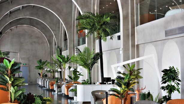 Um hall de hotel moderno com arcos curvos no teto, balcão de recepção, áreas de estar e diversas plantas em vasos.