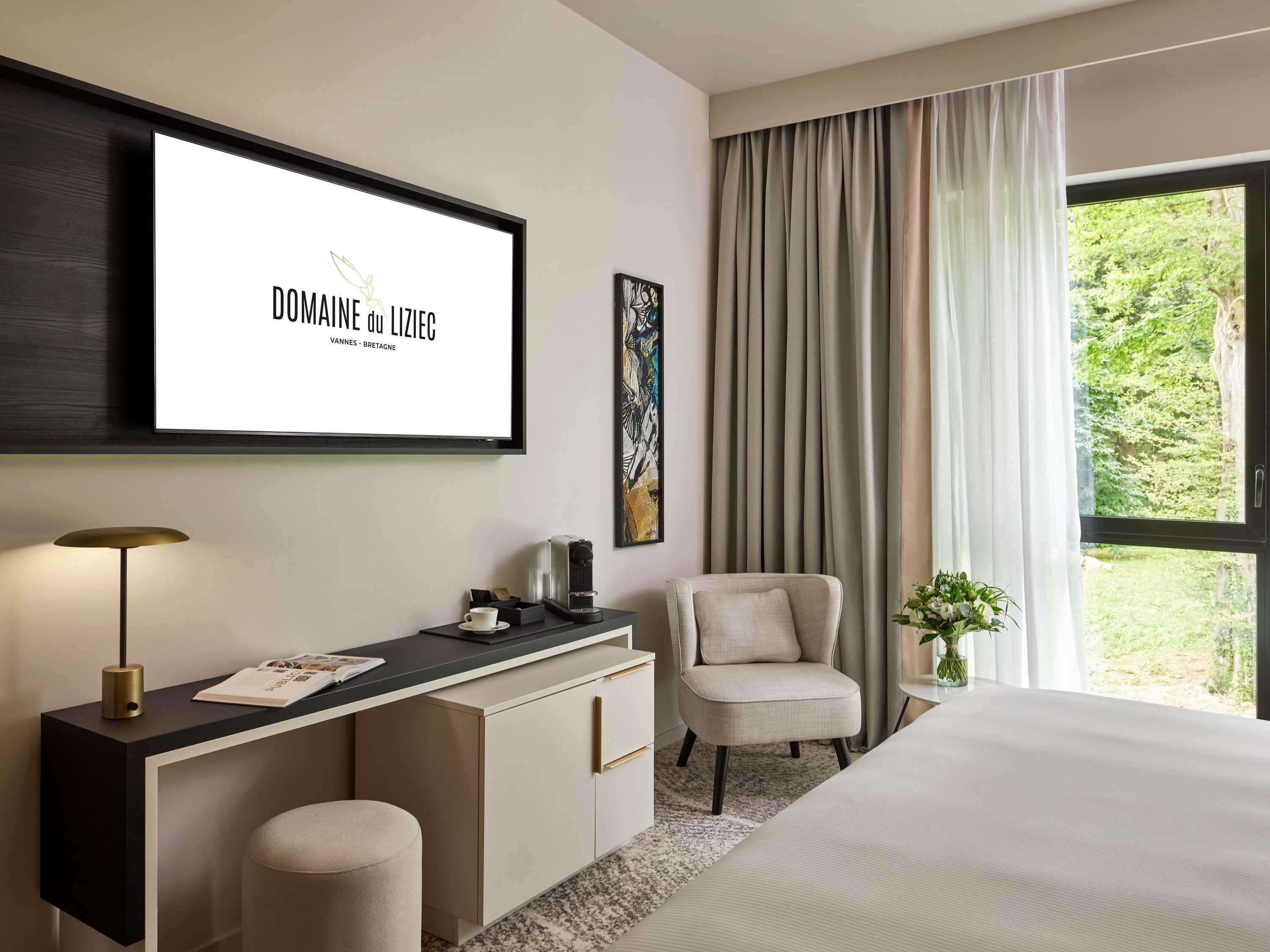 Quarto de hotel com cama, escrivaninha com luminária, cadeira, janela com cortinas e quadro na parede.