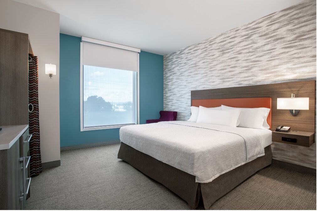 Quarto de hotel moderno com uma cama grande, uma mesa de cabeceira com abajur, uma janela com vista e um espaço para o banheiro.