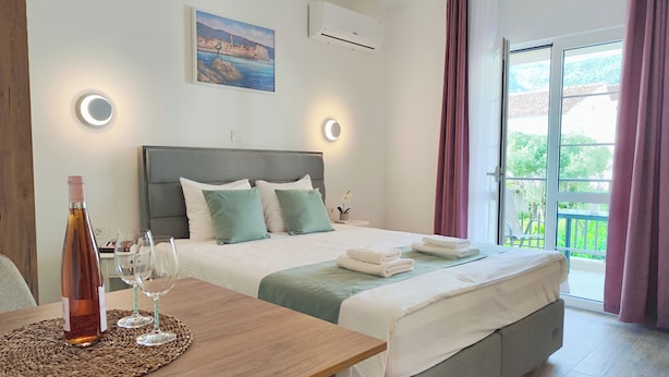 Quarto de hotel com cama, mesa de cabeceira com vinho e copos, um quadro na parede e uma varanda com cortinas.