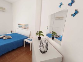 Quarto com duas camas, uma mesa pequena branca, uma planta em vaso, um espelho decorativo e ar condicionado fixado na parede.